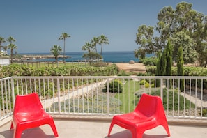 Uma varanda com duas cadeiras vermelhas, vista para o mar e um jardim com palmeiras.