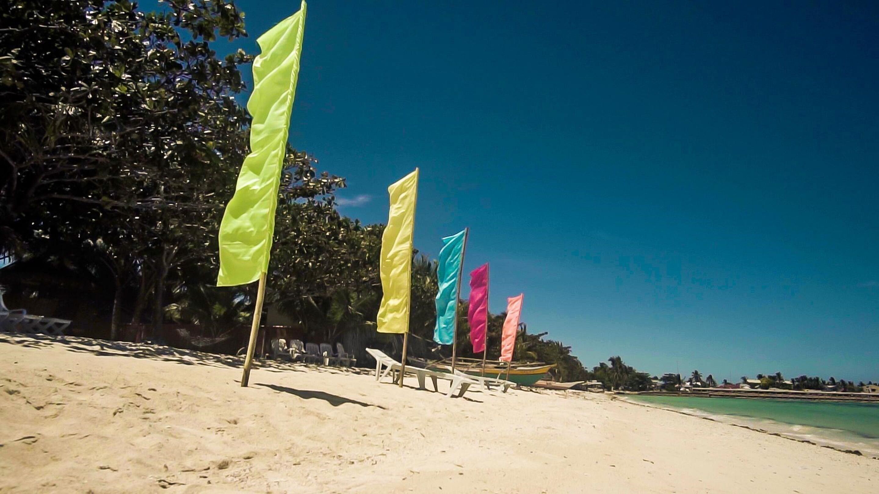 Uma praia com várias bandeiras coloridas, cadeiras de praia brancas e um céu azul claro.