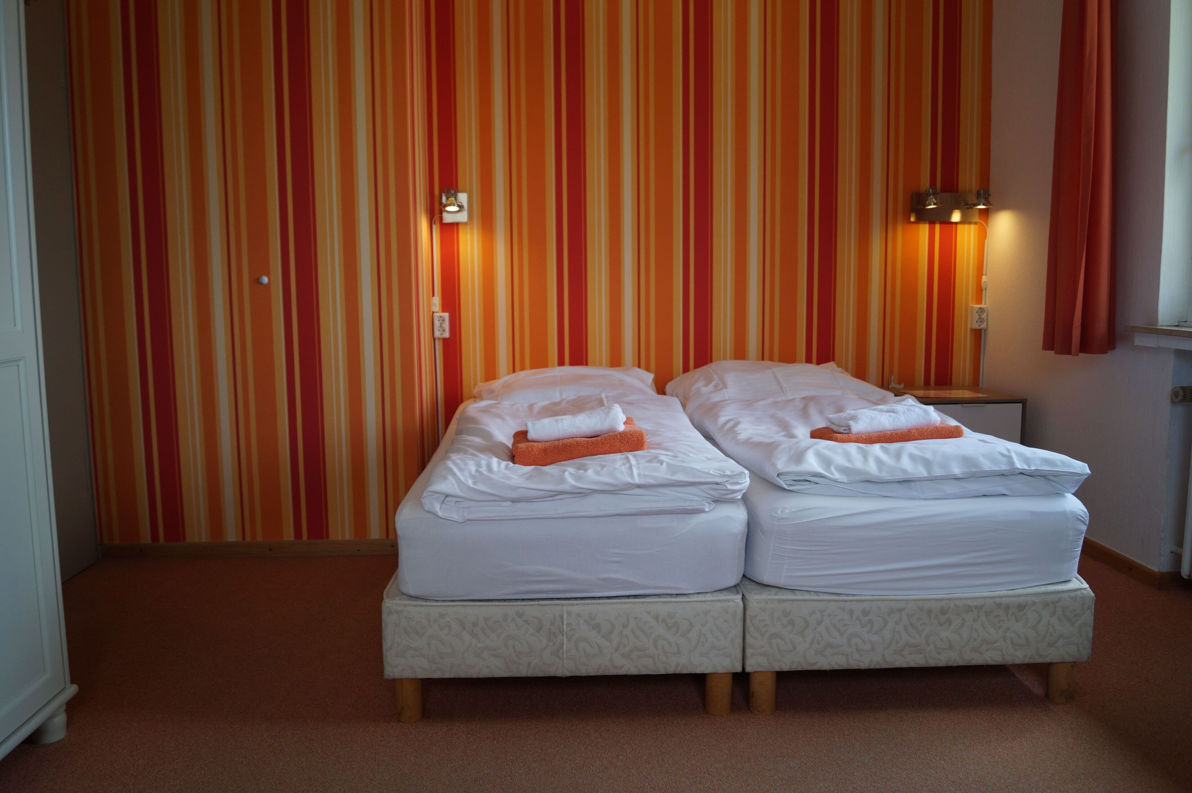 Uma cama de casal com lençóis brancos e toalhas laranja, uma parede listrada em laranja e vermelho, e uma mesa de cabeceira com uma luminária.