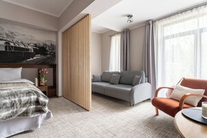 Quarto de hotel moderno com cama, sofá, poltrona, mesa pequena e uma grande janela com cortinas.