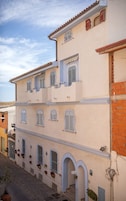 Um edifício de vários andares com fachada clara, diversas janelas com persianas e rua de paralelepípedos.