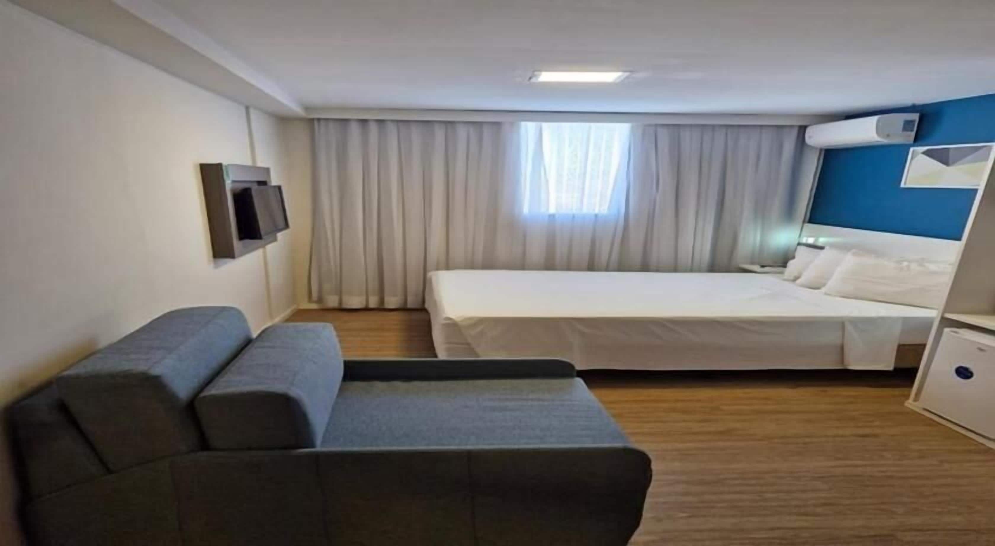 Quarto de hotel com cama, sofá, televisão e janela com cortinas.