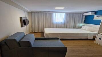 Quarto de hotel com cama, sofá, televisão e janela com cortinas.