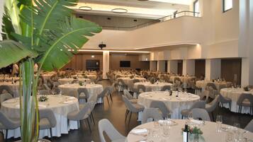 Uma ampla sala de banquetes com várias mesas redondas montadas para refeições, rodeada por cadeiras.