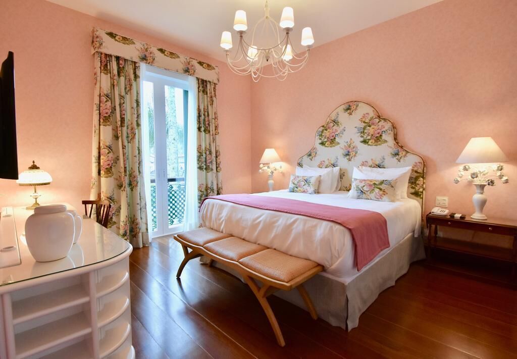 Quarto com cama, luminárias de cabeceira, um banco, escrivaninha e lustre.