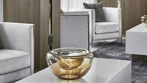 Quarto de hotel moderno com uma janela grande, um sofá, uma poltrona e uma mesa de centro com uma tigela decorativa.