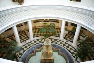 Um hall de hotel com piso quadriculado, uma fonte central e colunas brancas.