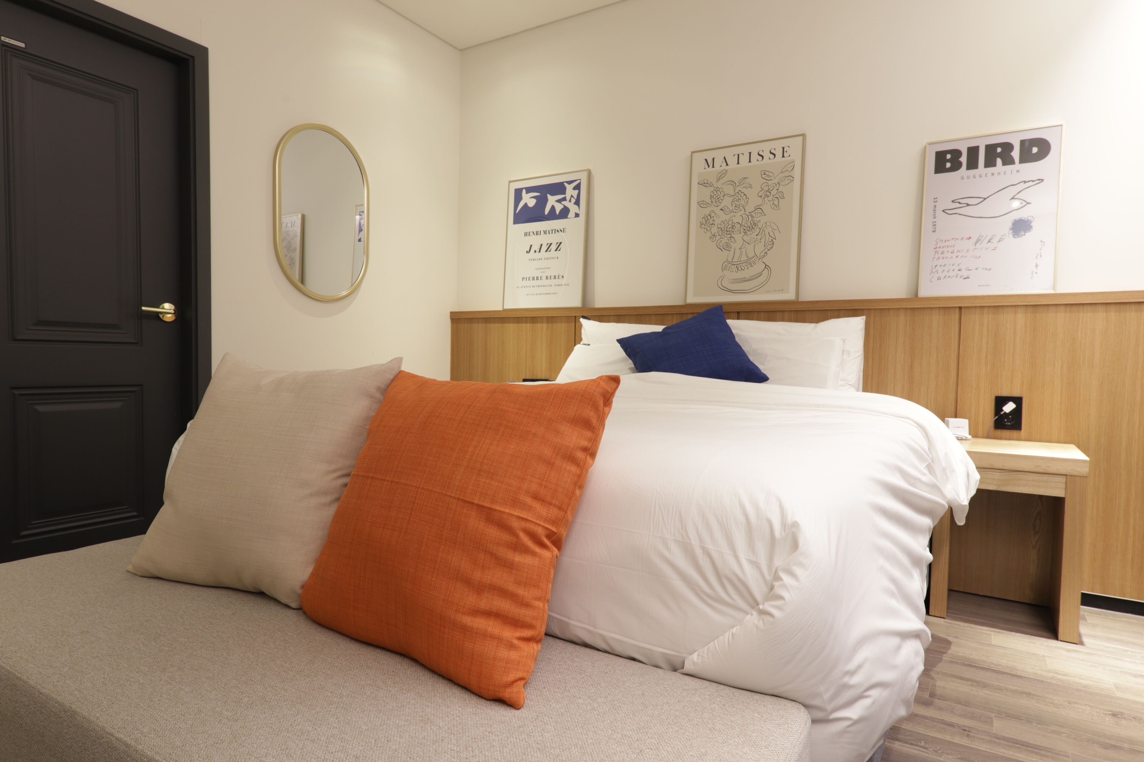 Cama bem arrumada com roupa de cama branca e azul, um travesseiro laranja e uma mesa de cabeceira de madeira com abajur.