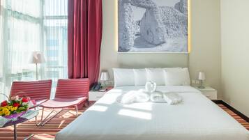 Quarto de hotel com uma cama grande, duas cadeiras vermelhas, uma escrivaninha com abajur e uma janela com cortinas.
