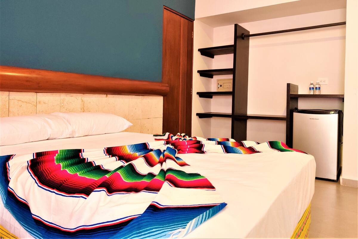 Quarto de hotel com cama, cabeceira de madeira, prateleira na parede com garrafas de água e um pequeno frigorífico.