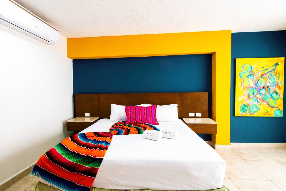 Quarto de hotel com uma cama, mesa de cabeceira, ar condicionado fixado na parede e um quadro colorido.
