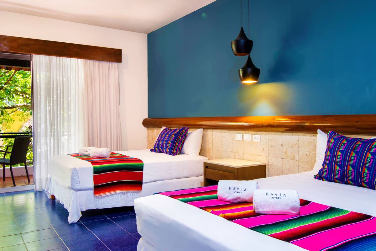 Quarto de hotel com duas camas, cada uma com um cobertor colorido e uma almofada com etiqueta de nome. Há uma cabeceira de madeira, uma mesa de cabeceira e uma janela com cortinas.