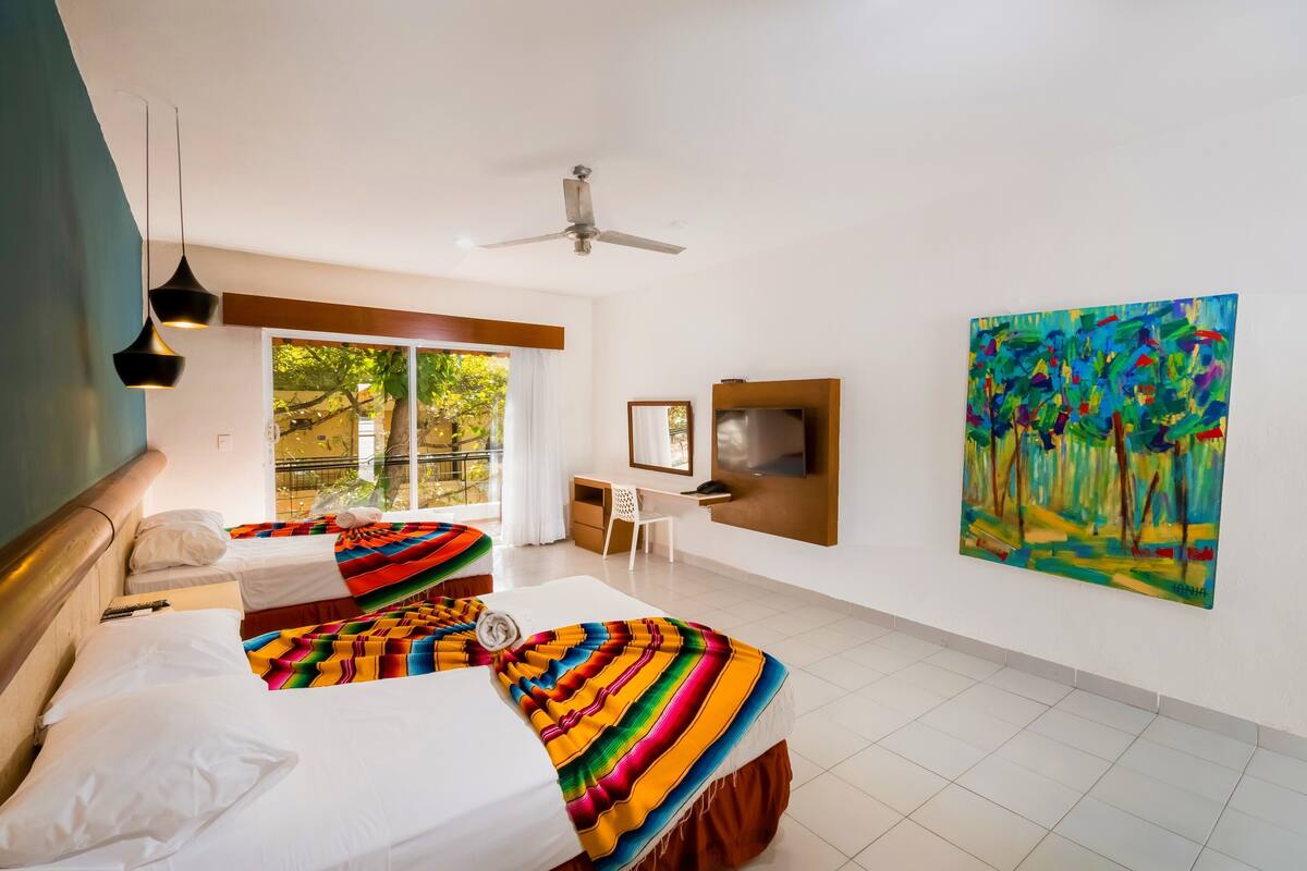 Quarto de hotel com duas camas, uma escrivaninha, um ventilador de teto, uma janela grande e um quadro na parede.