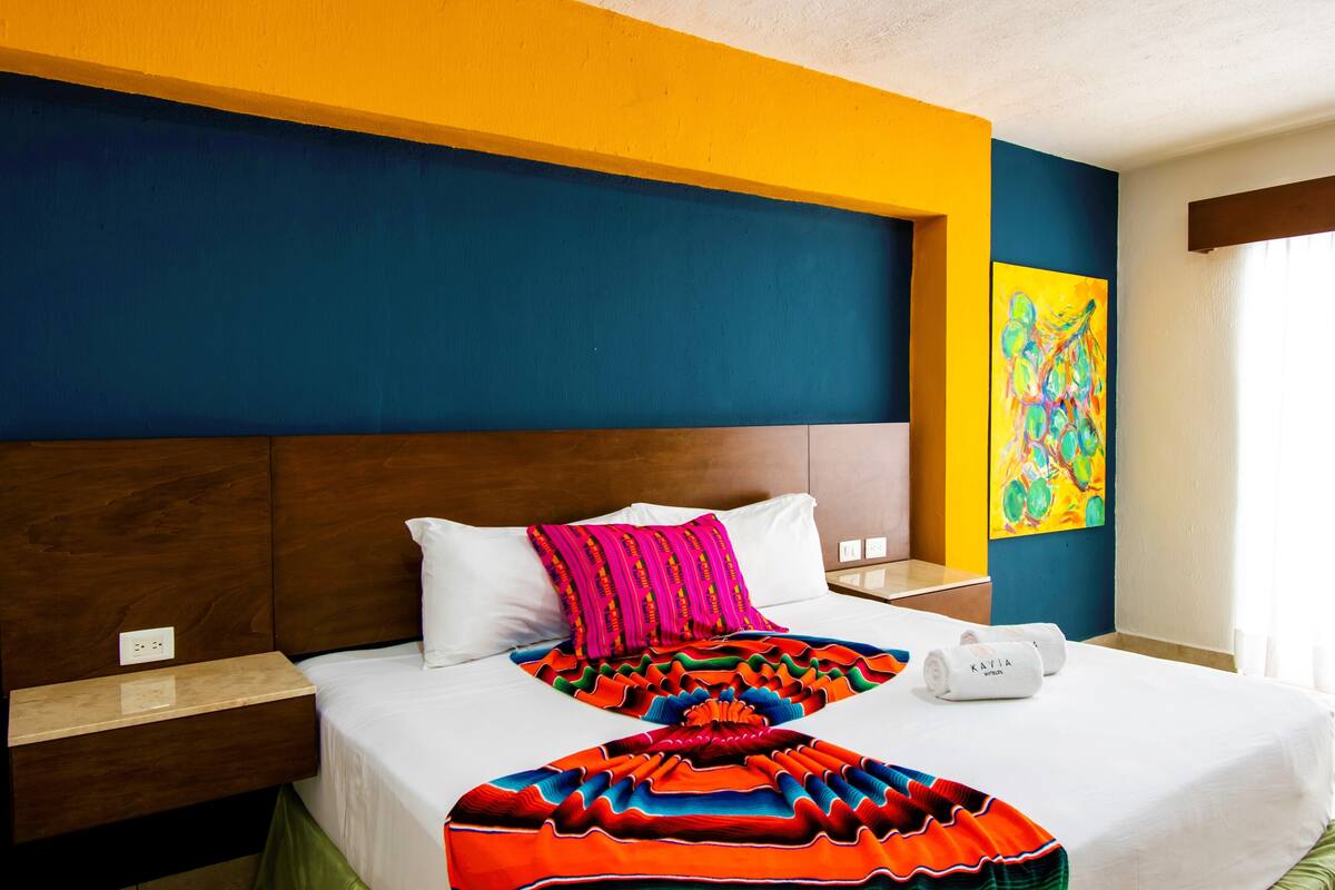 Quarto de hotel com uma cama grande, cabeceira de madeira, manta colorida e um quadro na parede.