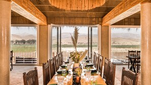 Uma sala de jantar com uma mesa comprida posta para refeição, grandes janelas com vista para o deserto e teto de madeira com detalhes entalhados.