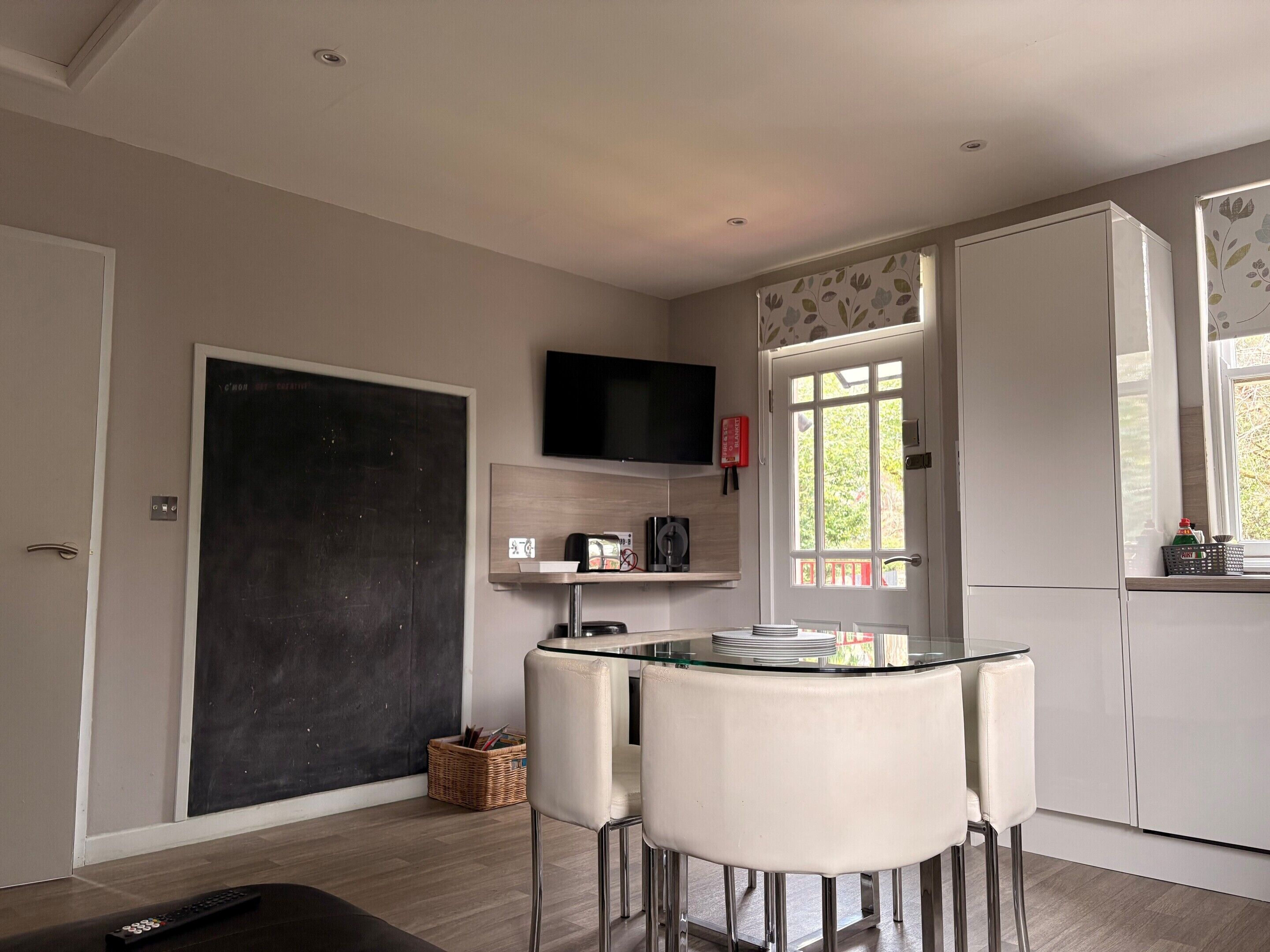 Uma cozinha moderna com uma mesa de vidro, cadeiras brancas, um quadro negro e uma televisão sobre o balcão.