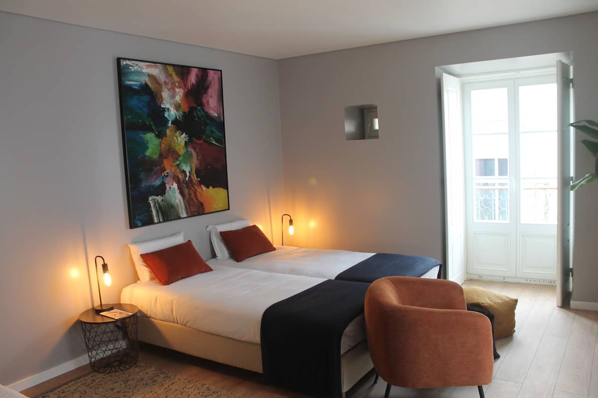 Quarto moderno com duas camas, uma pintura abstrata grande, uma mesa de cabeceira com abajur e vista para o exterior através de uma porta.