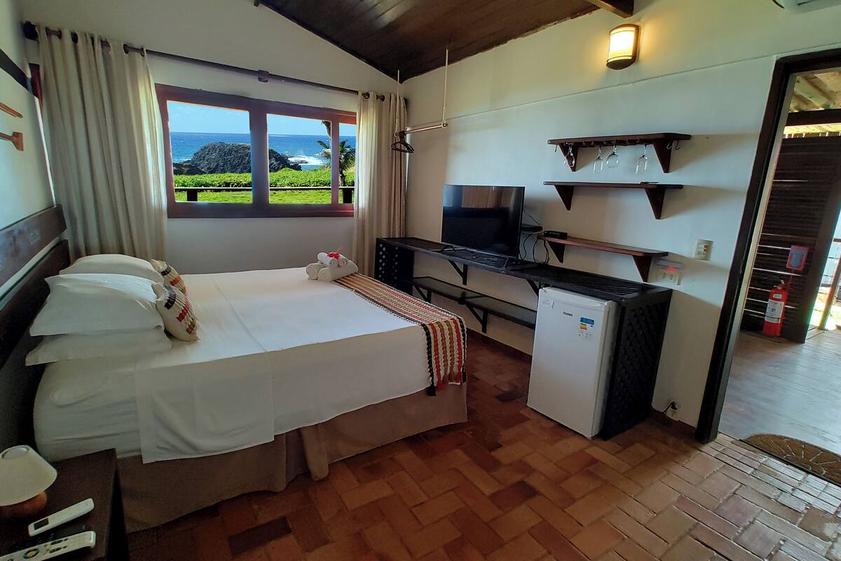 Quarto com cama, escrivaninha com televisão, mini-frigorífico e vista para o oceano.