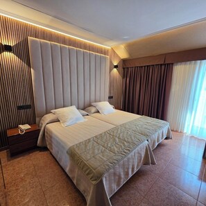 Uma cama de casal com cabeceira, uma mesa de cabeceira com abajur e uma janela com cortinas.