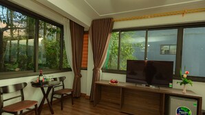 Quarto com televisão, mini-frigorífico, mesa de jantar com cadeiras e janelas amplas com cortinas.