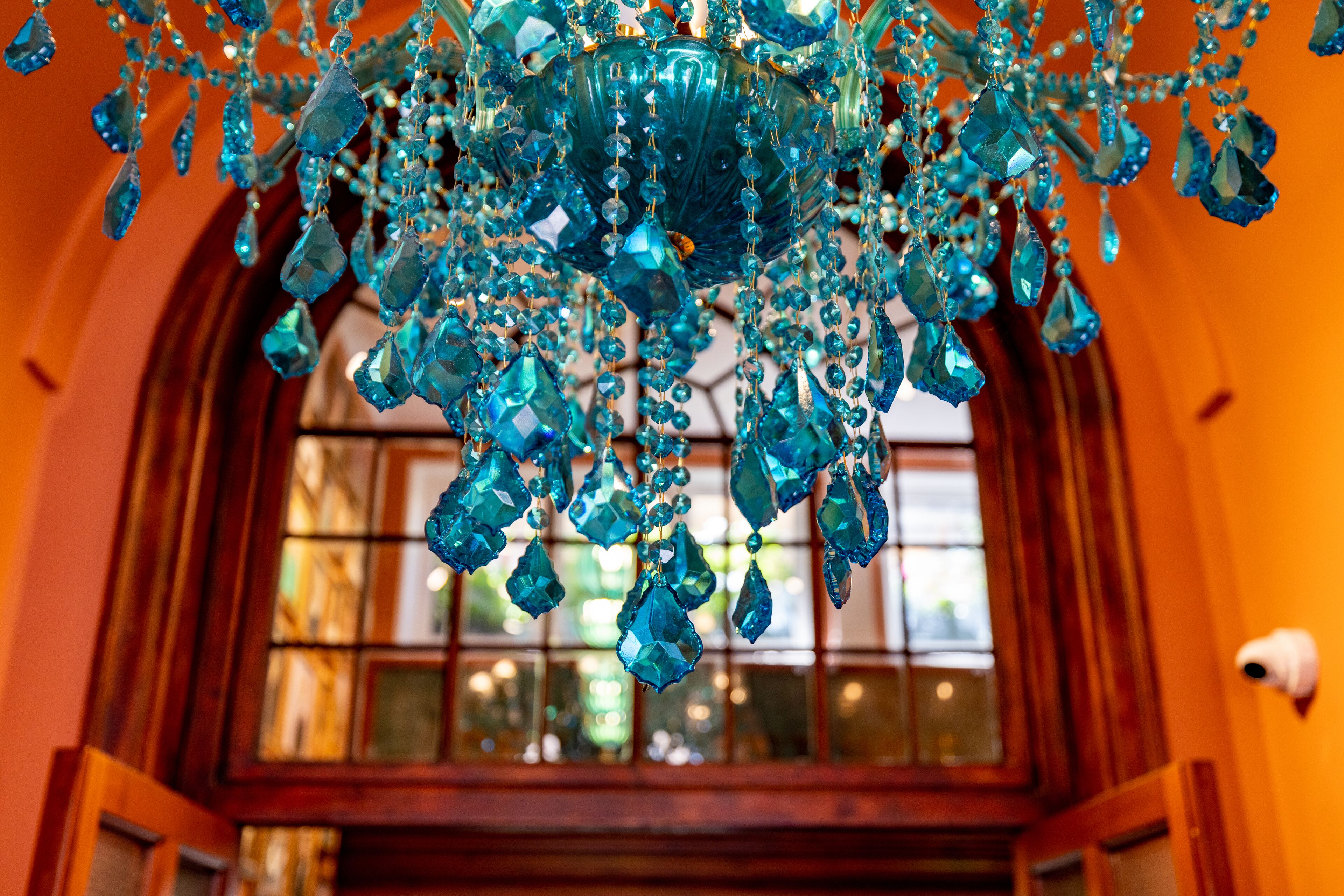 Um candelabro com detalhes em cristal azul pendurado em um ambiente com arquitetura em madeira e uma janela ao fundo.