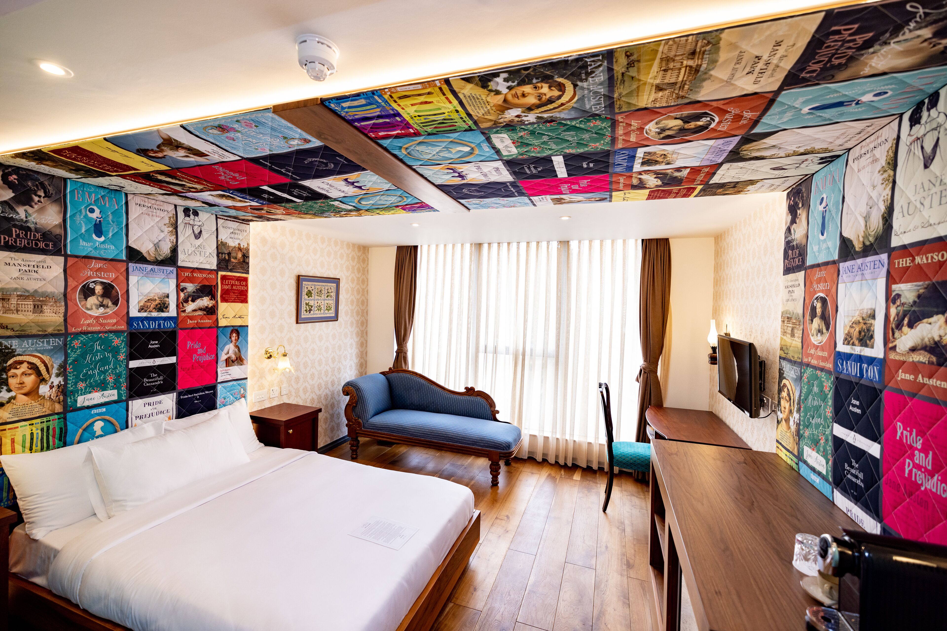 Quarto de hotel com uma cama grande, uma poltrona azul, uma escrivaninha e uma parede decorada com cartazes de livros.