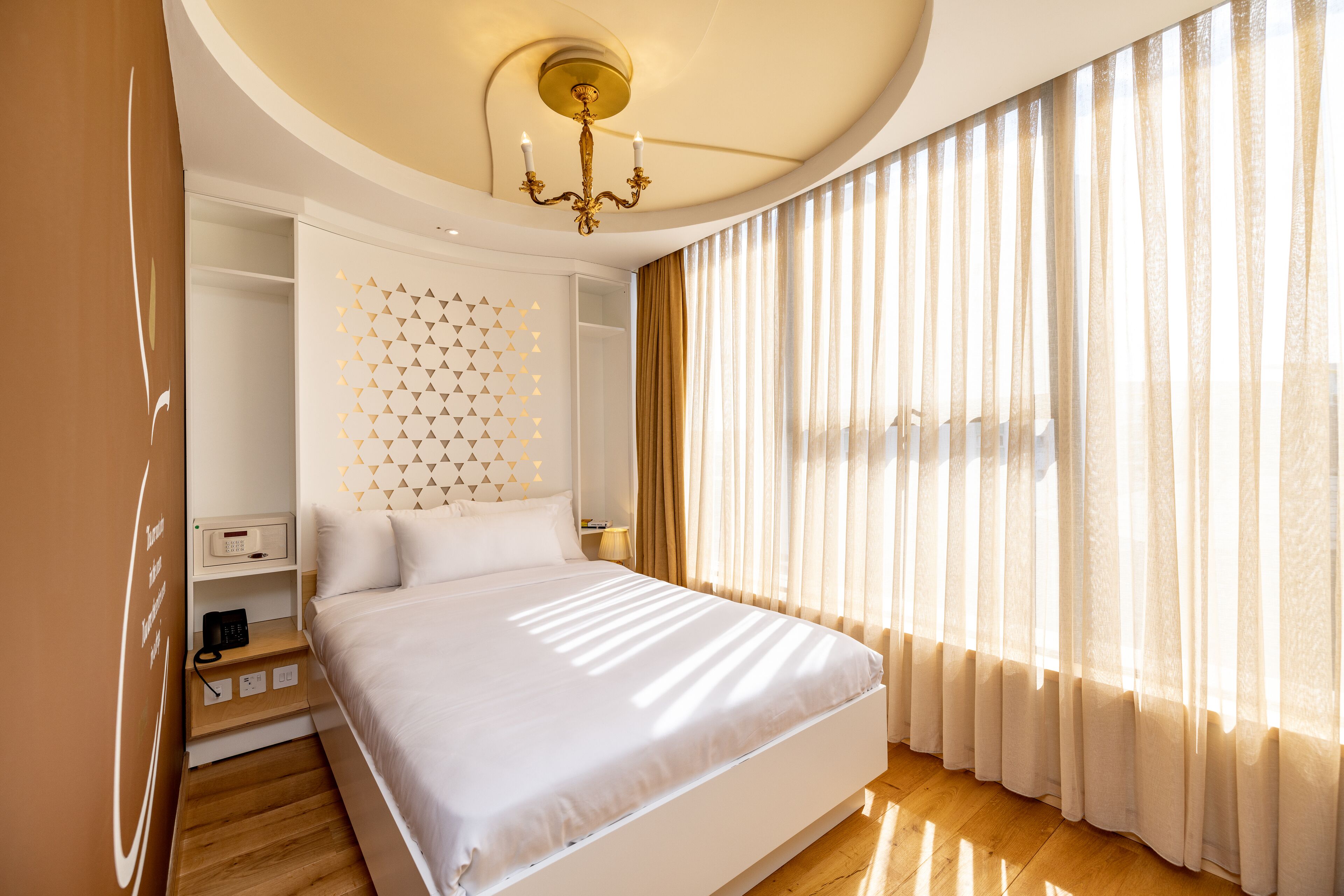 Quarto de hotel moderno com uma cama grande, uma mesa de cabeceira, um relógio fixado na parede e uma janela com cortinas leves.