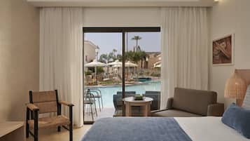 Quarto de hotel com cama, uma cadeira, uma mesinha e vista para uma piscina e palmeiras.