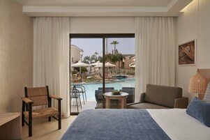 Quarto de hotel com cama, uma cadeira, uma mesinha e vista para uma piscina e palmeiras.