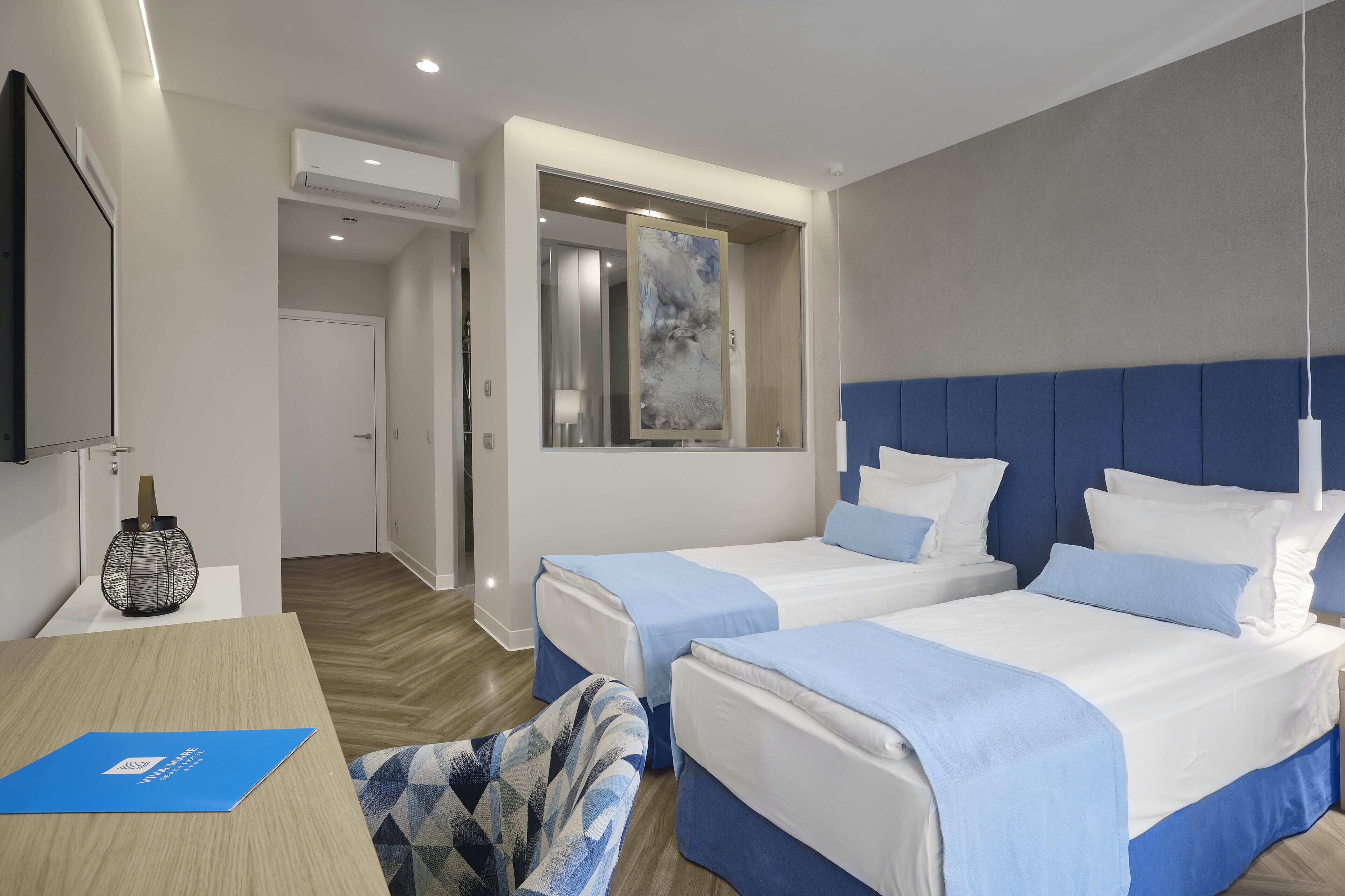 Quarto de hotel com duas camas, uma mesa e uma cadeira. Há uma janela grande com vista para o exterior.
