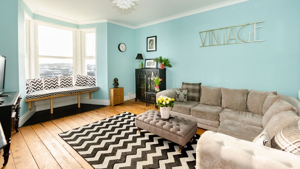 Uma sala de estar com parede azul, sofá bege, tapete preto e branco e um banco com almofadas estampadas.