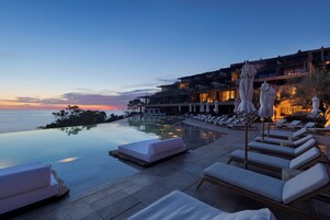 Piscina infinita com espreguiçadeiras e guarda-sóis, com vista para o pôr do sol.