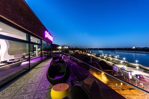 Um edifício moderno com a placa 'Pizza', mesas ao ar livre com cadeiras pretas e amarelas distintas, e vista para um corpo d'água e luzes da cidade ao entardecer.