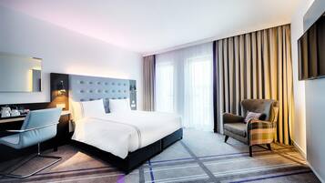 Quarto de hotel com cama, escrivaninha com cadeira, televisão e uma janela grande com cortinas.