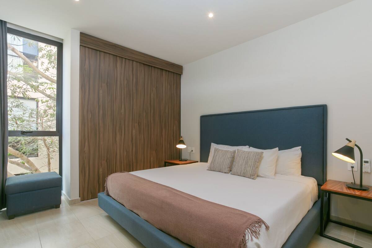 Um quarto moderno com uma cama grande, cabeceira de madeira, mesa de cabeceira com abajur e vista para o exterior por uma janela ampla.