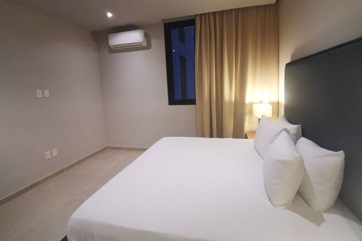Quarto de hotel com cama, janela com cortinas, ar-condicionado e mesa de cabeceira com abajur.