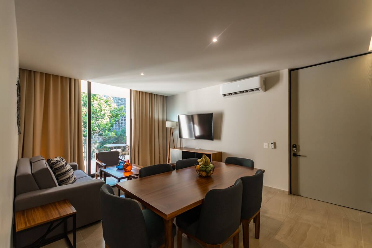 Uma sala de estar moderna com um sofá, uma mesa de jantar e cadeiras. Uma televisão de tela plana está fixada na parede.