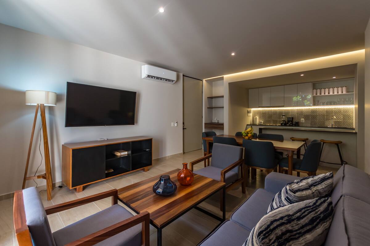 Uma sala de estar moderna com televisão de tela plana, mesa de centro de madeira, sofá e área de jantar com cadeiras e mesa.