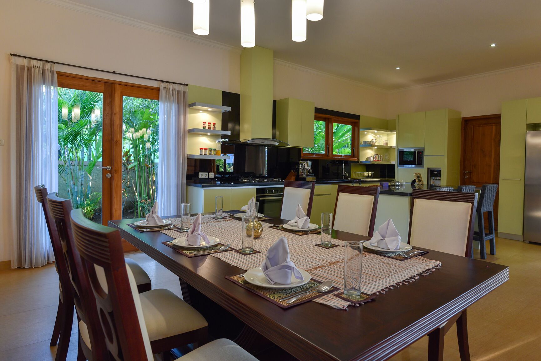 Uma área de jantar com mesa posta para quatro pessoas, cozinha ao fundo e vista para o verde pela janela.