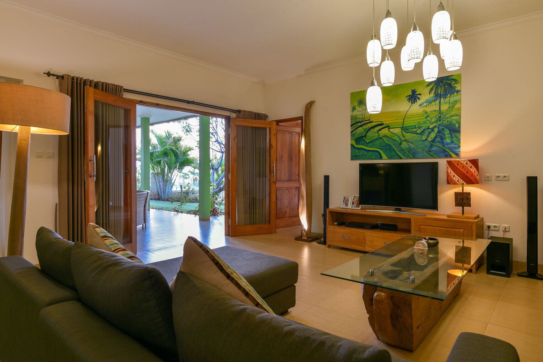 Uma sala de estar moderna com sofá, TV, mesa de centro de vidro e um quadro de paisagem tropical.