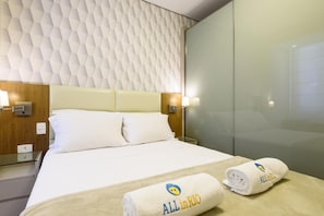 Quarto de hotel com cama, duas almofadas, mesa de cabeceira com abajur e duas toalhas com a inscrição 'ALL in RIO'.