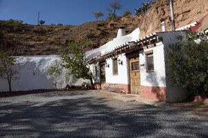 Um edifício branco com porta e janelas de madeira, rodeado por cascalho e vegetação rala.