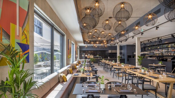 Restaurante moderno com mesas comunitárias longas, área de bar e janelas amplas com vista para a rua.