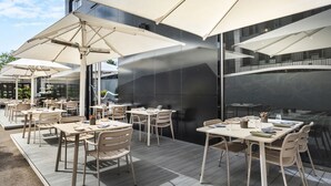 Uma área de refeições ao ar livre com várias mesas e cadeiras, grandes guarda-sóis e uma fachada de prédio moderno ao fundo.