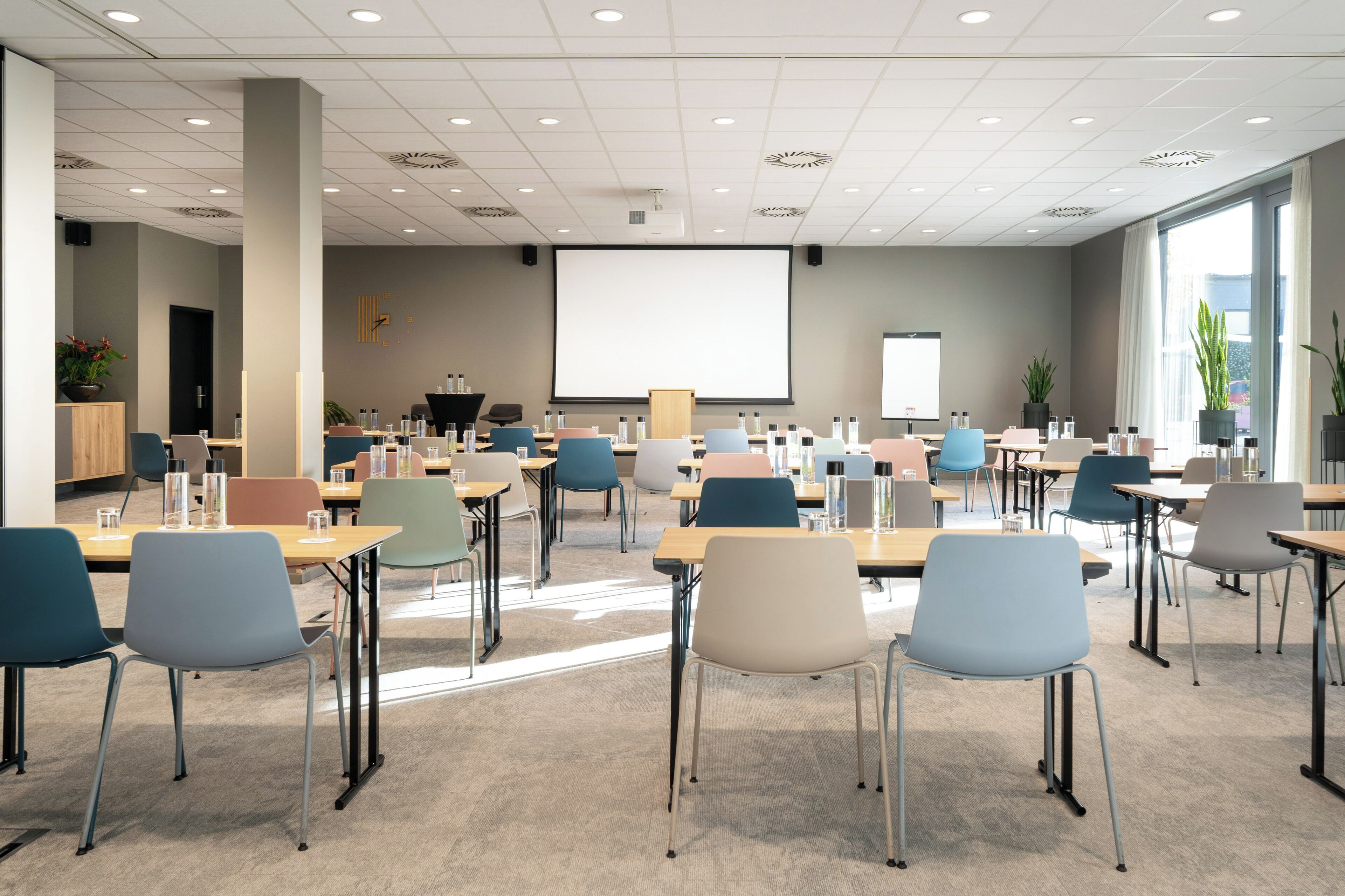 Uma sala de conferências com várias mesas e cadeiras, uma tela de projetor e uma janela grande.