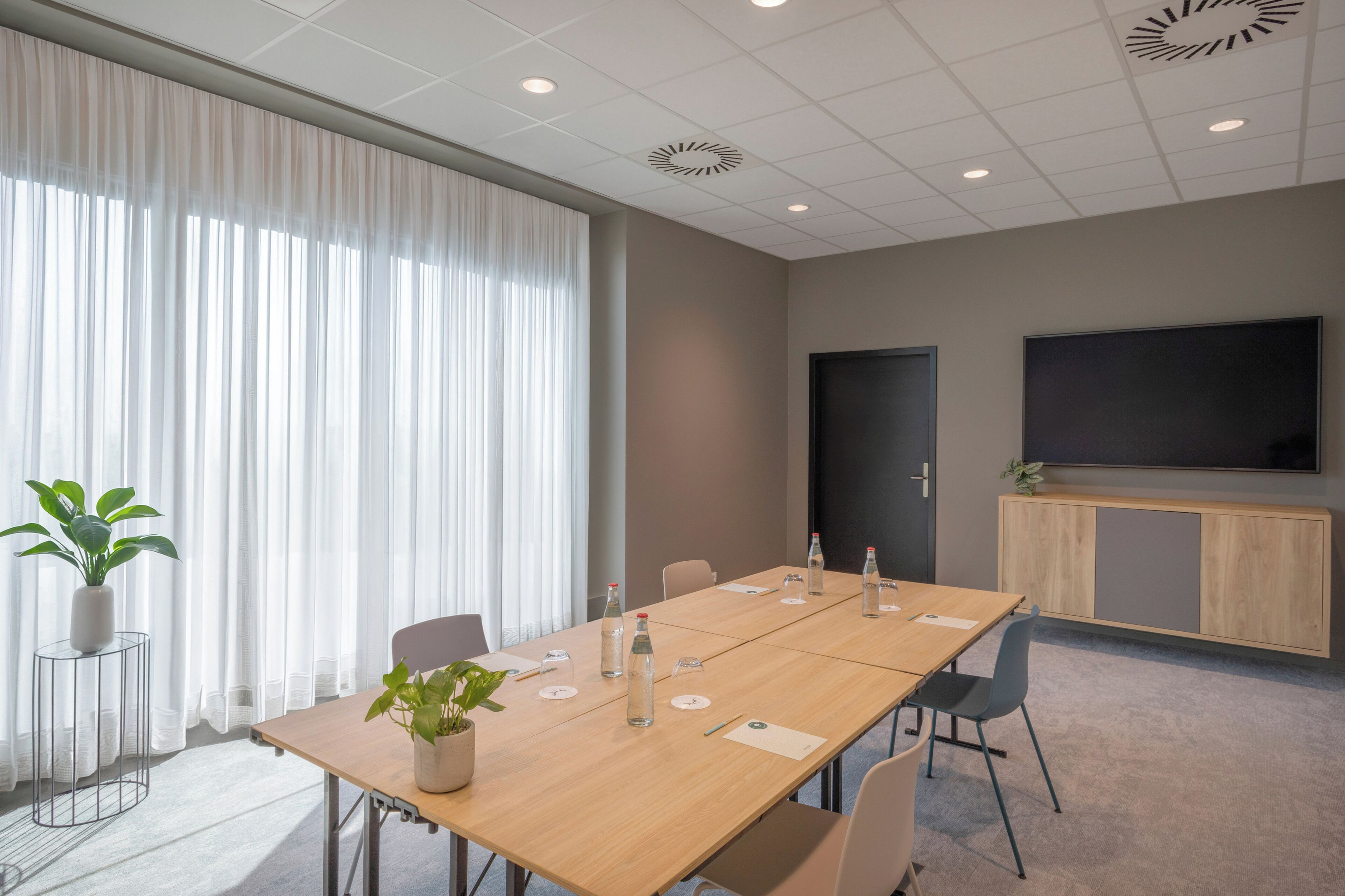 Uma sala de conferência com uma mesa comprida, cadeiras, uma planta e uma televisão sobre um suporte.