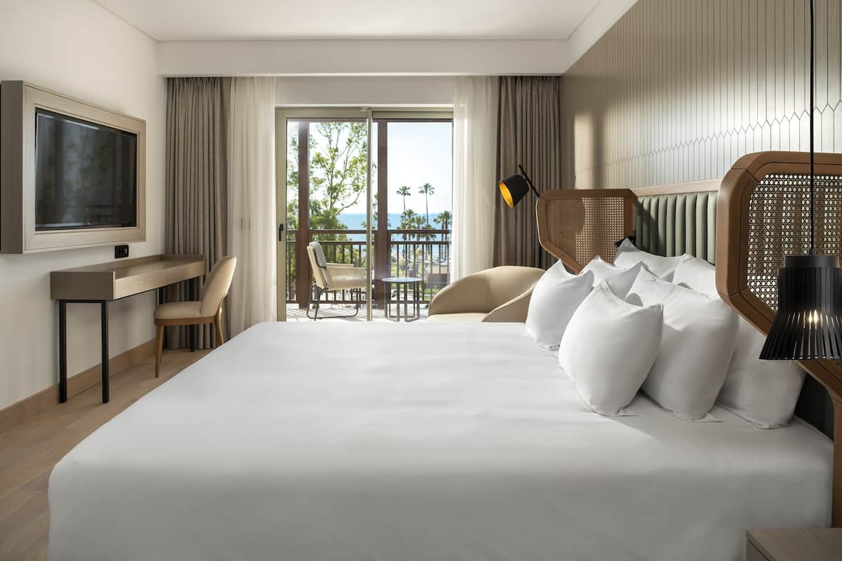 Quarto de hotel com uma cama grande, uma escrivaninha, uma cadeira e uma televisão. Há uma varanda com vista para palmeiras e o oceano.