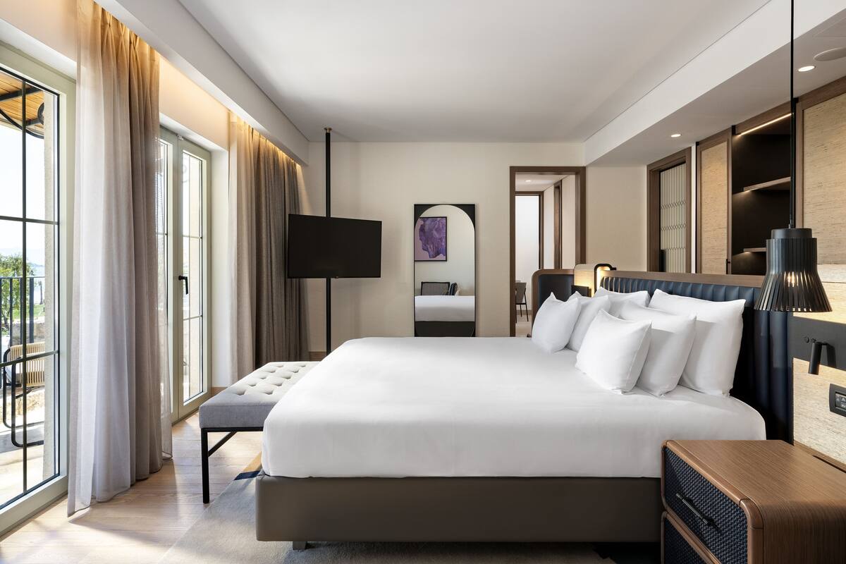 Quarto de hotel moderno com uma cama grande, televisão de tela plana, área de estar e vista para o exterior.