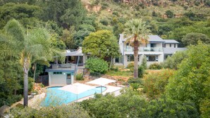 Uma casa de dois andares com piscina, rodeada por vegetação exuberante e um morro rochoso.