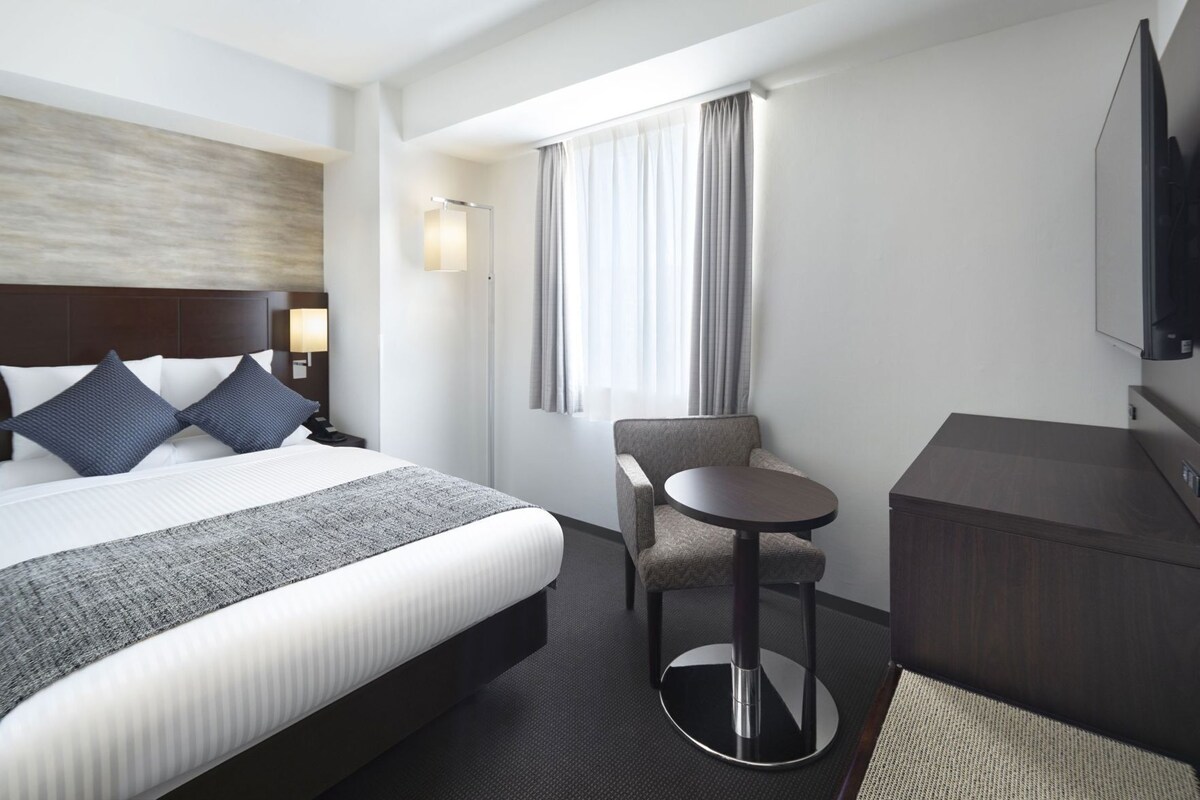 Quarto de hotel com cama, mesa pequena redonda, cadeira, televisão e janela com cortinas.